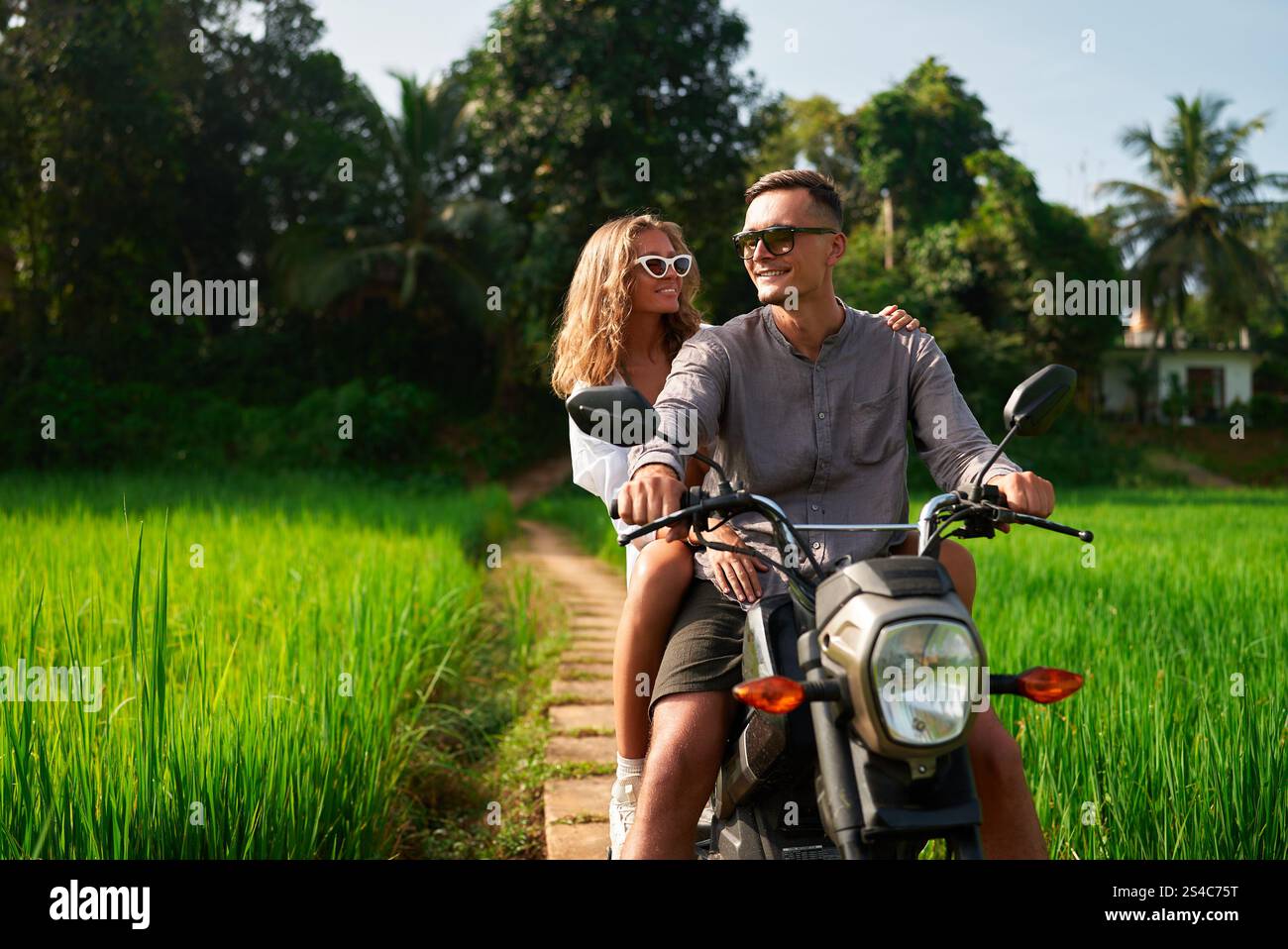 Couple rides motorbike through green rice field path. Man drives, woman ...