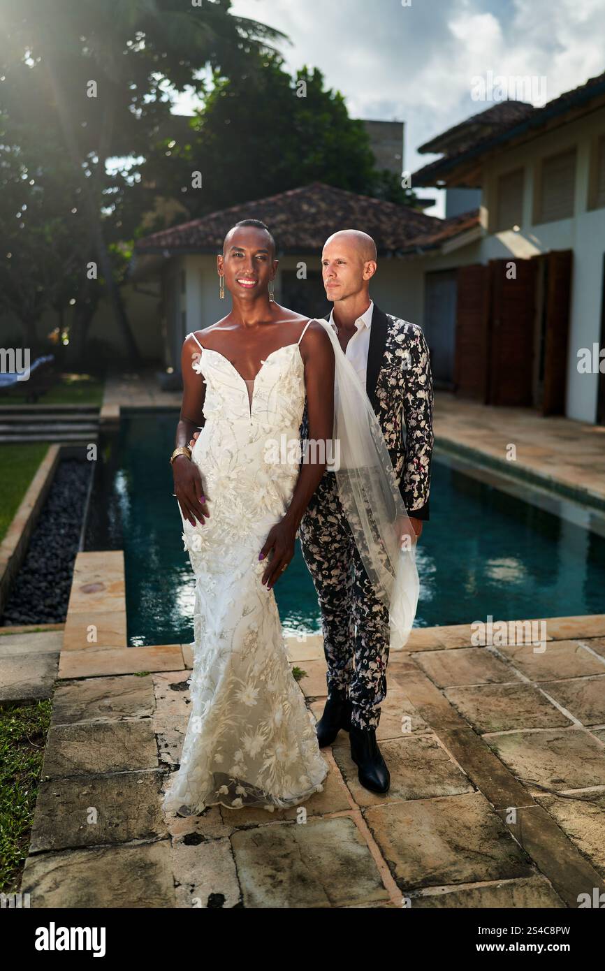 Gender fluid person in white dress with LGBTQ groom in suit near pool ...