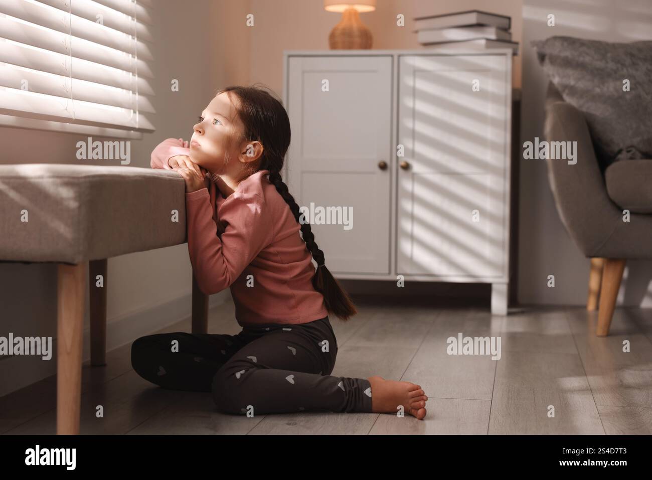 Orphanage concept. Sad girl and looking through window blinds indoors ...