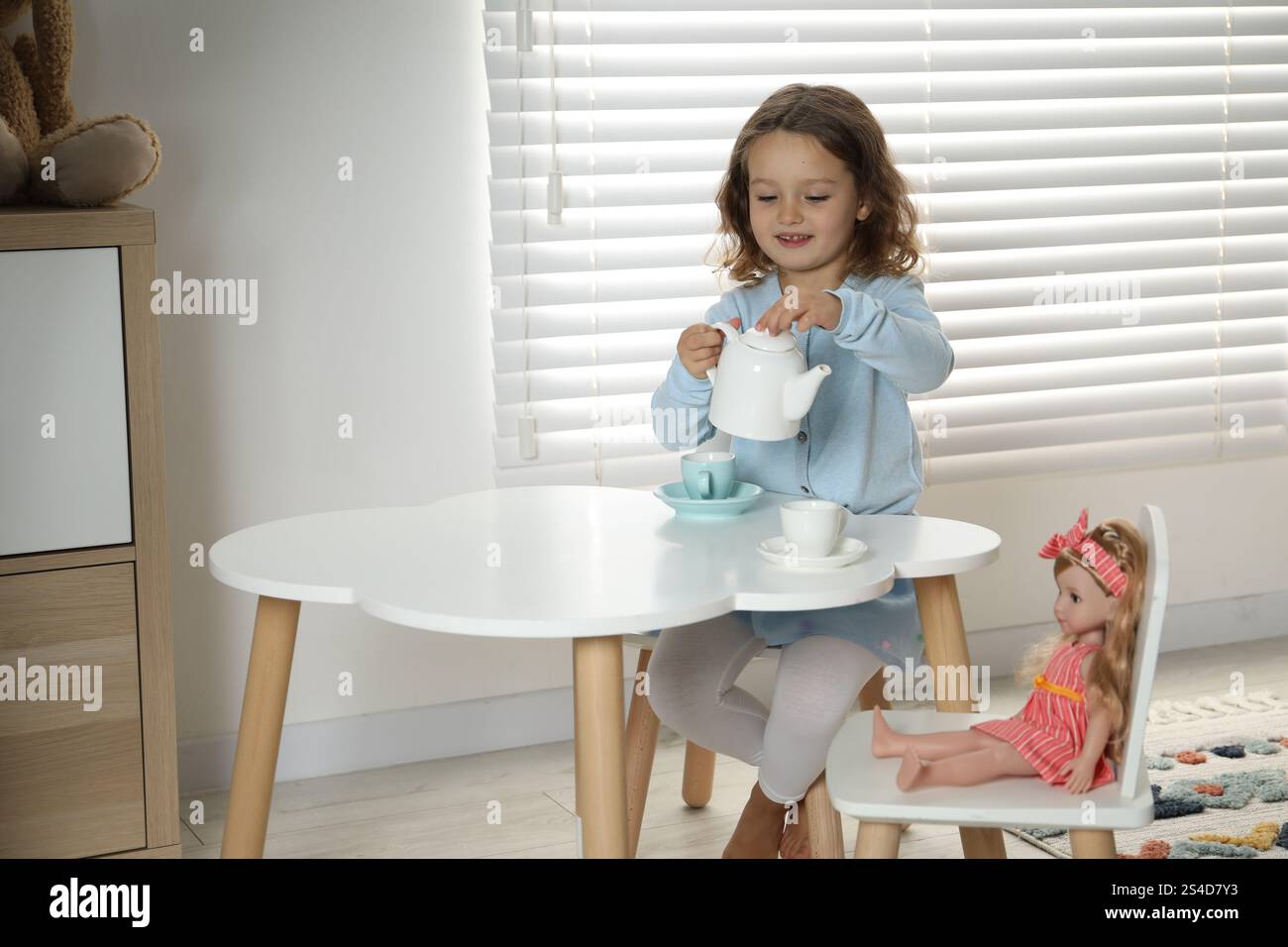 Cute little girl playing tea party with doll at white table in room ...