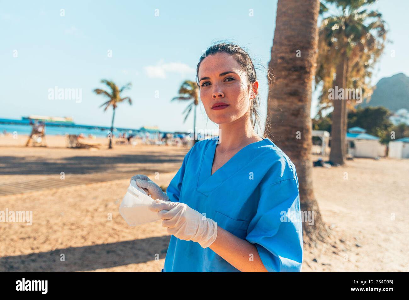 A nurse in a blue scrub suit is standing on a beach holding a face mask ...