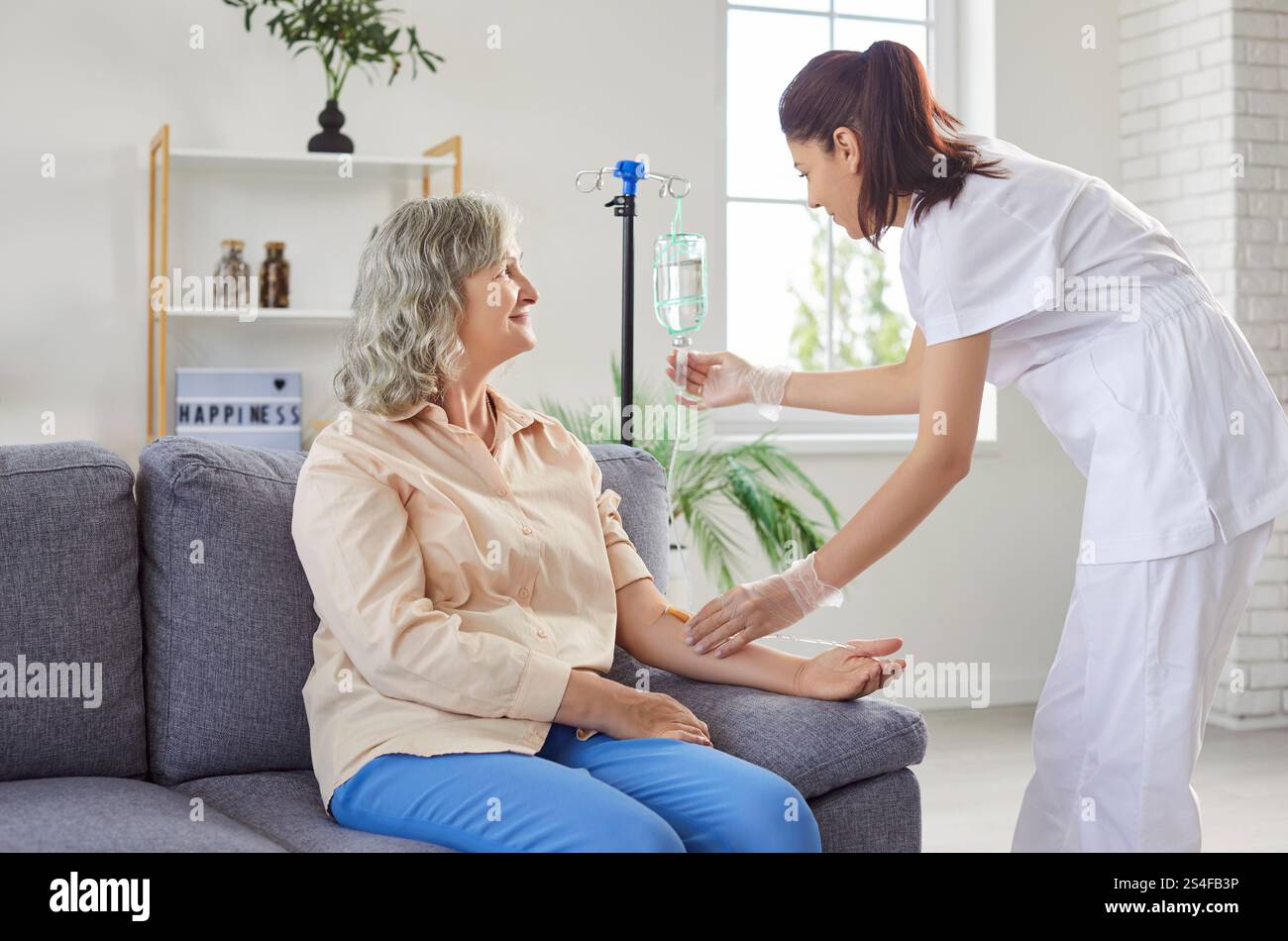 Young nurse helping elderly woman sitting on sofa to receive IV drip ...