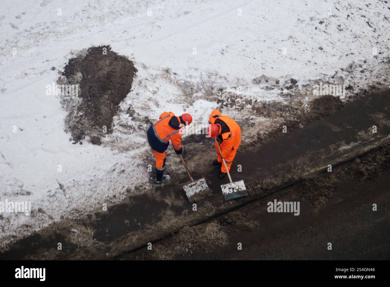 Workers in orange safety suits shoveling snow and dirt on a winter road ...