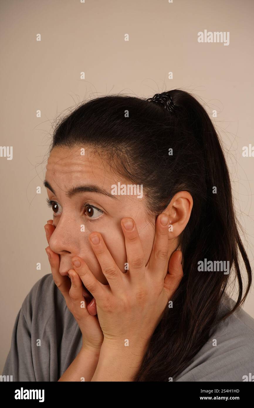 A young woman holds her hands over her face, her eyes wide open and her ...