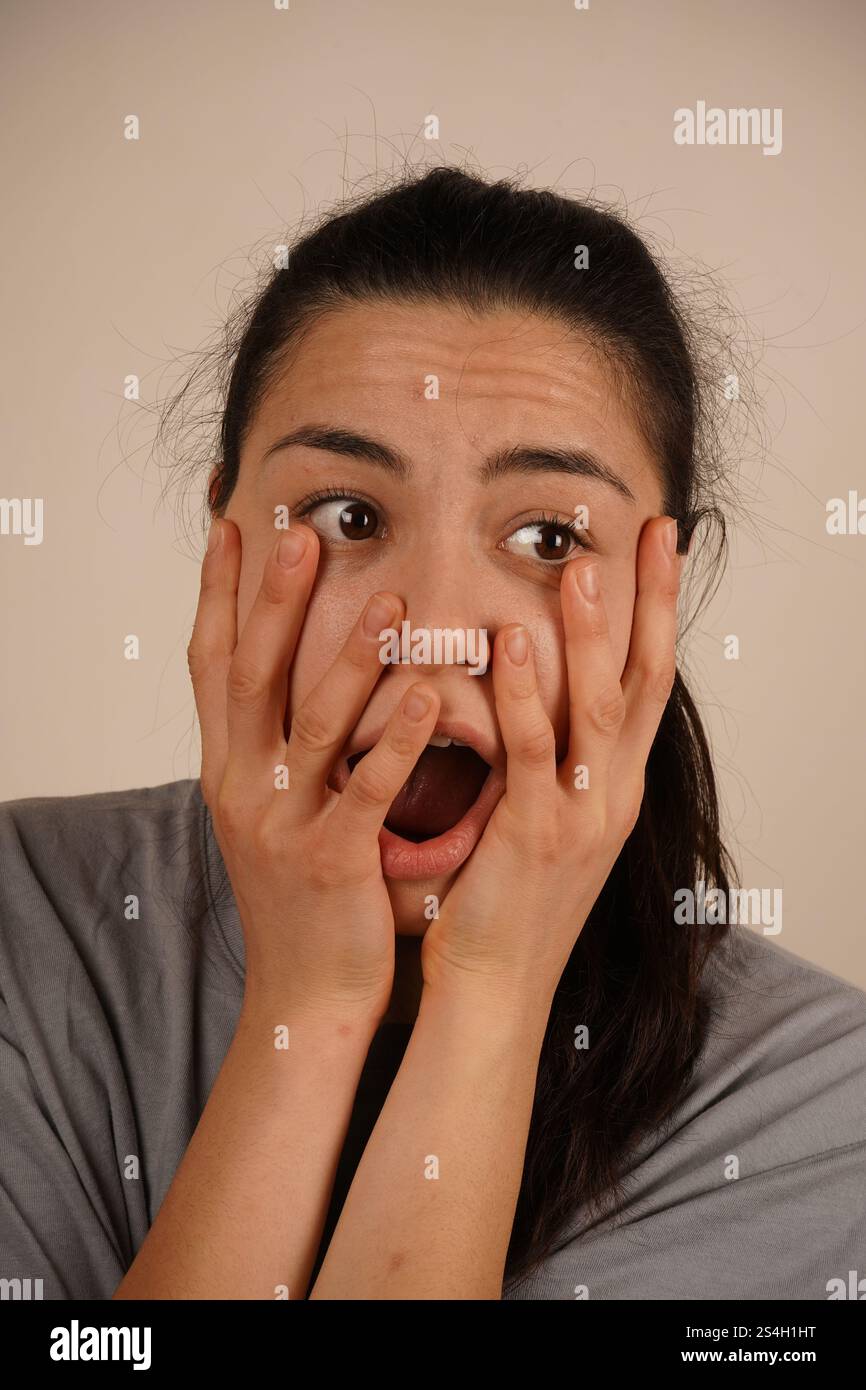 A young woman holds her hands over her face, her eyes wide open and her ...