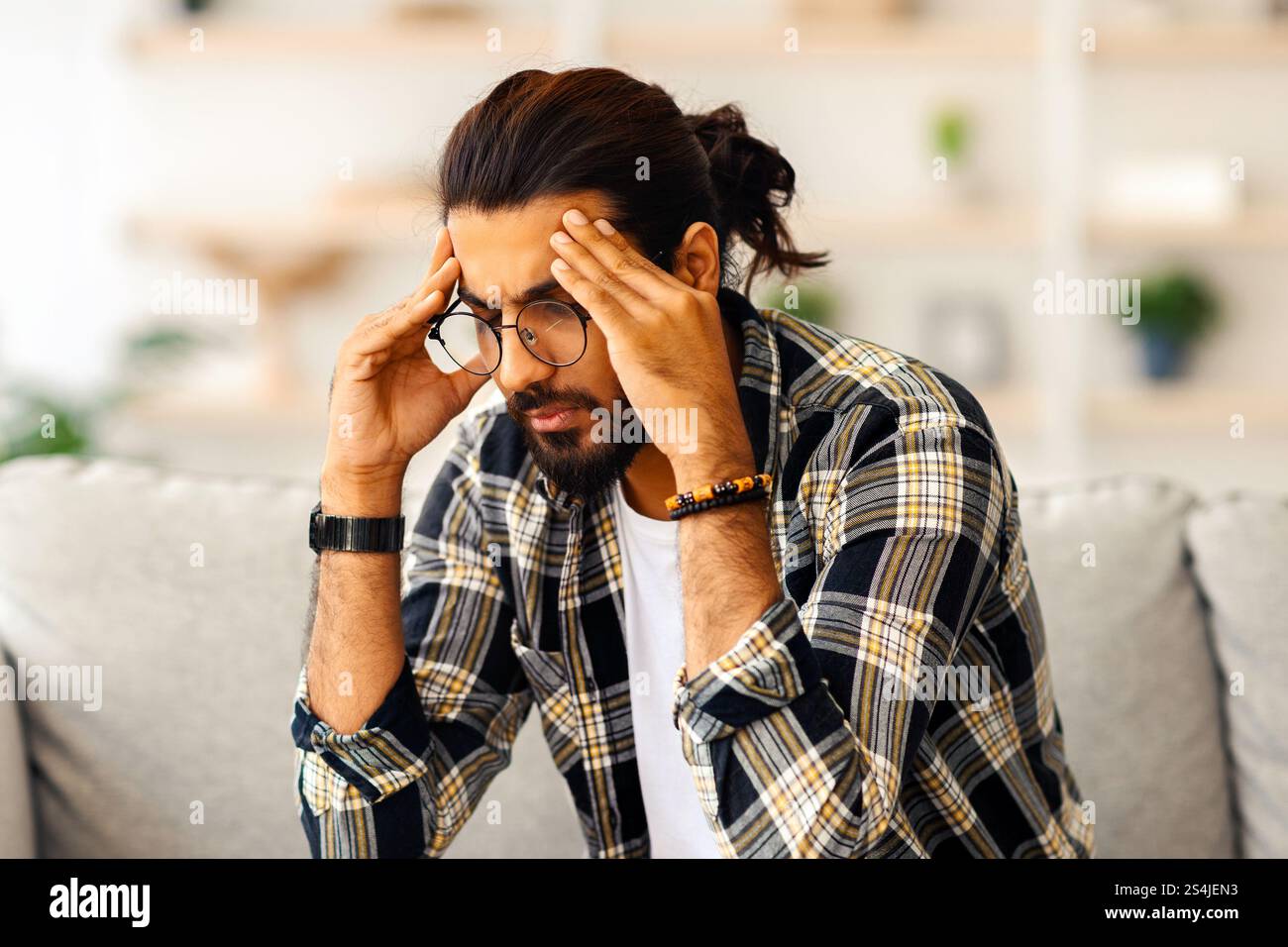 Indian guy sitting on couch at home, touching head Stock Photo - Alamy