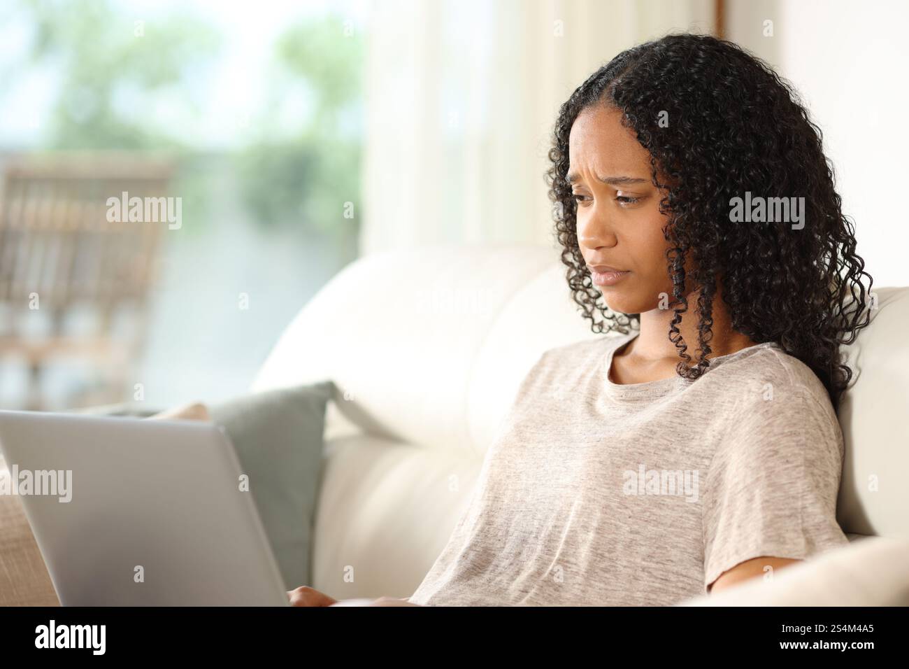 Serious black woman using laptop sitting on a couch in a house interior ...