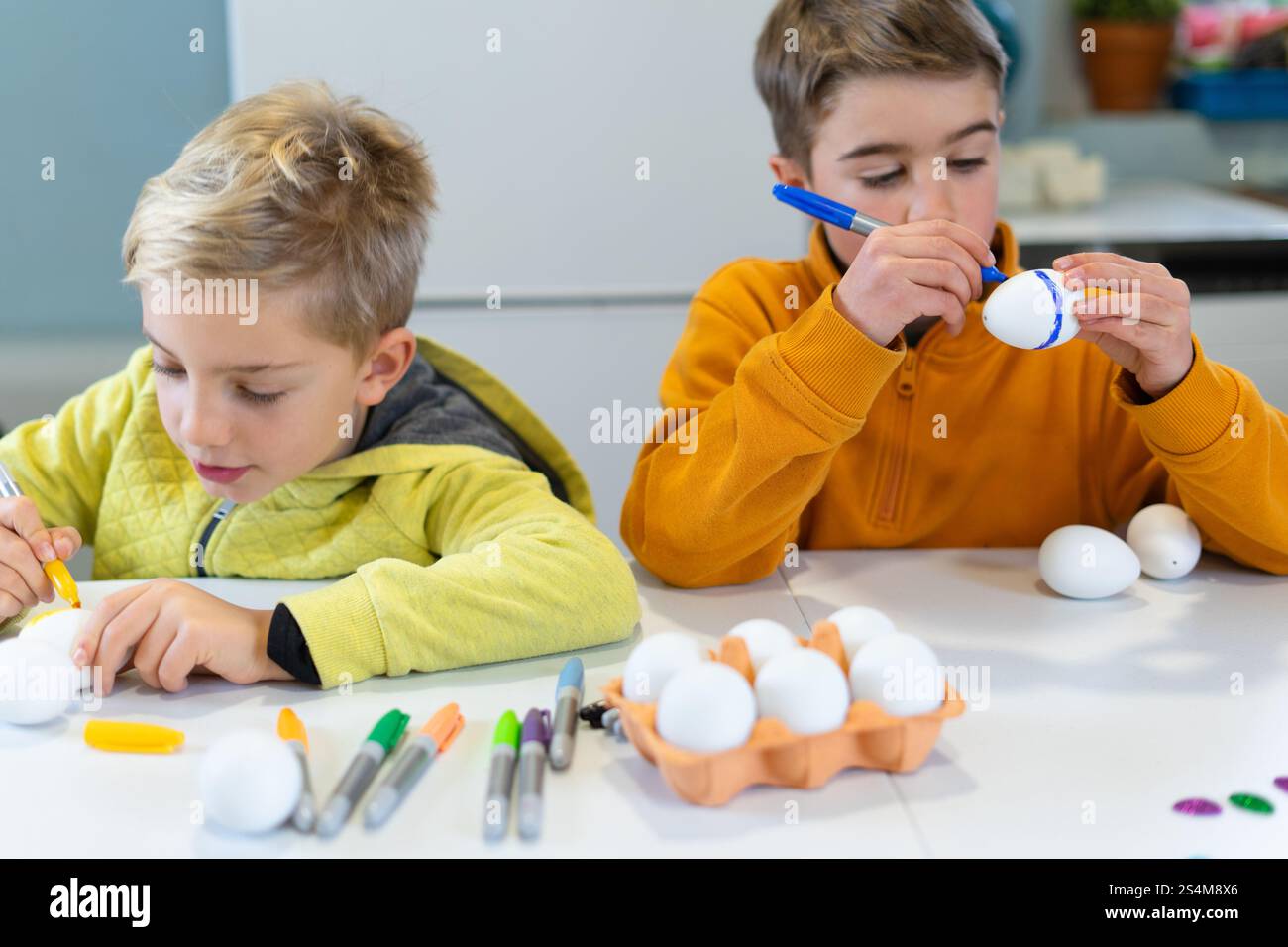 Two brothers are decorating white eggs for easter using colorful markers, sitting at a white ...