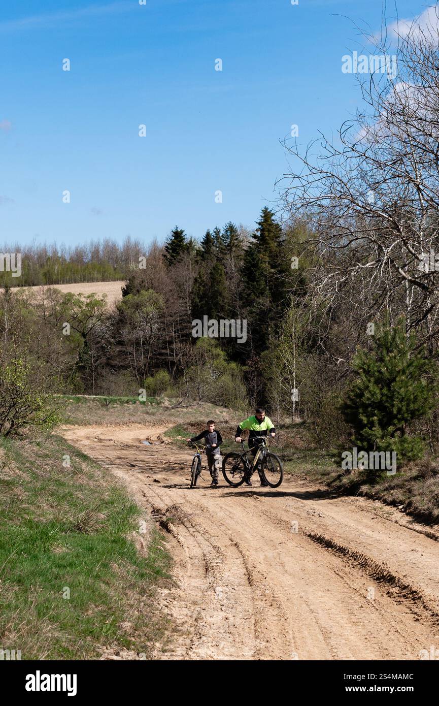 A man and a boy enjoy a bike ride down a sandy path. Trees line the ...