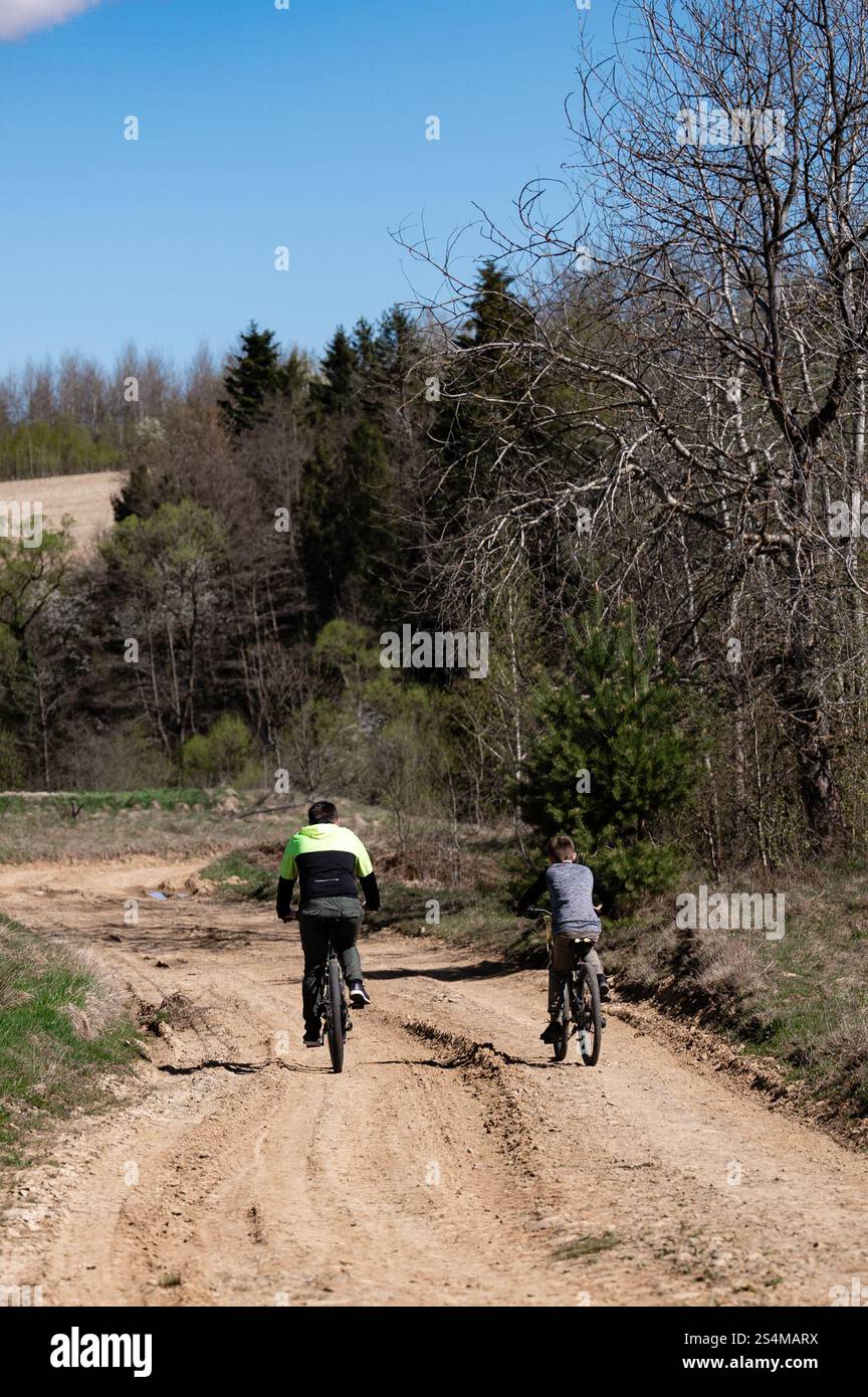Two people bike along a sandy path. Trees and foliage surround the path ...