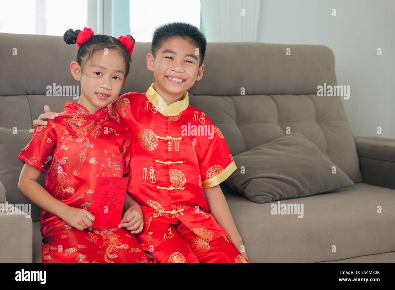 Smiling children in red cheongsam sit on a sofa, holding angpao red ...