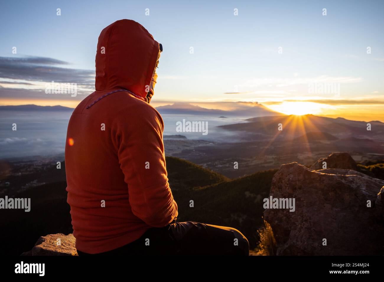 Back view of unrecognizable man sitting atop a mountain, watching the ...