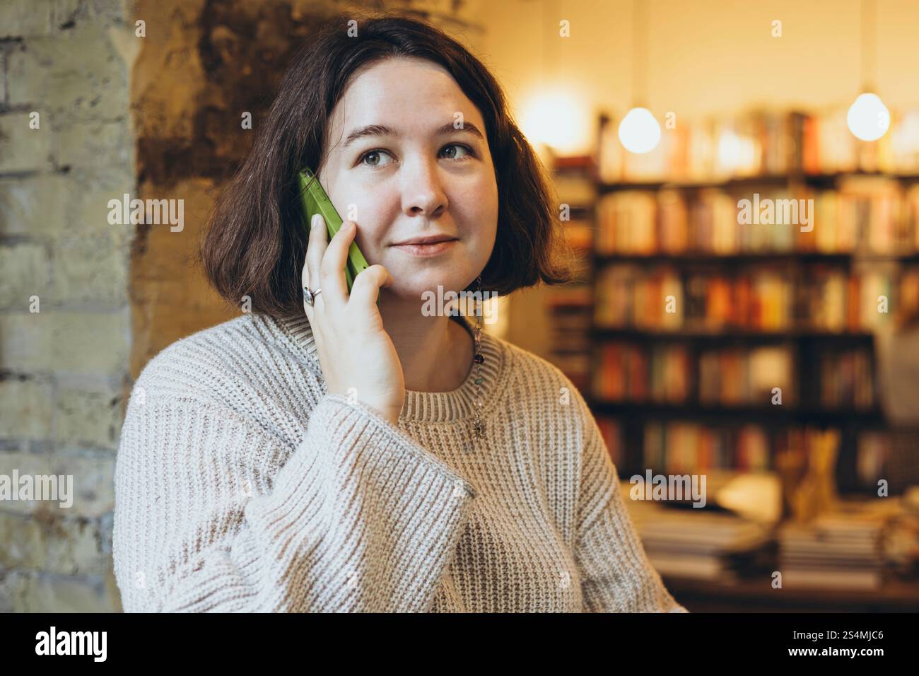 A young woman in a cream sweater smiles while talking on her green smartphone inside a warm ...