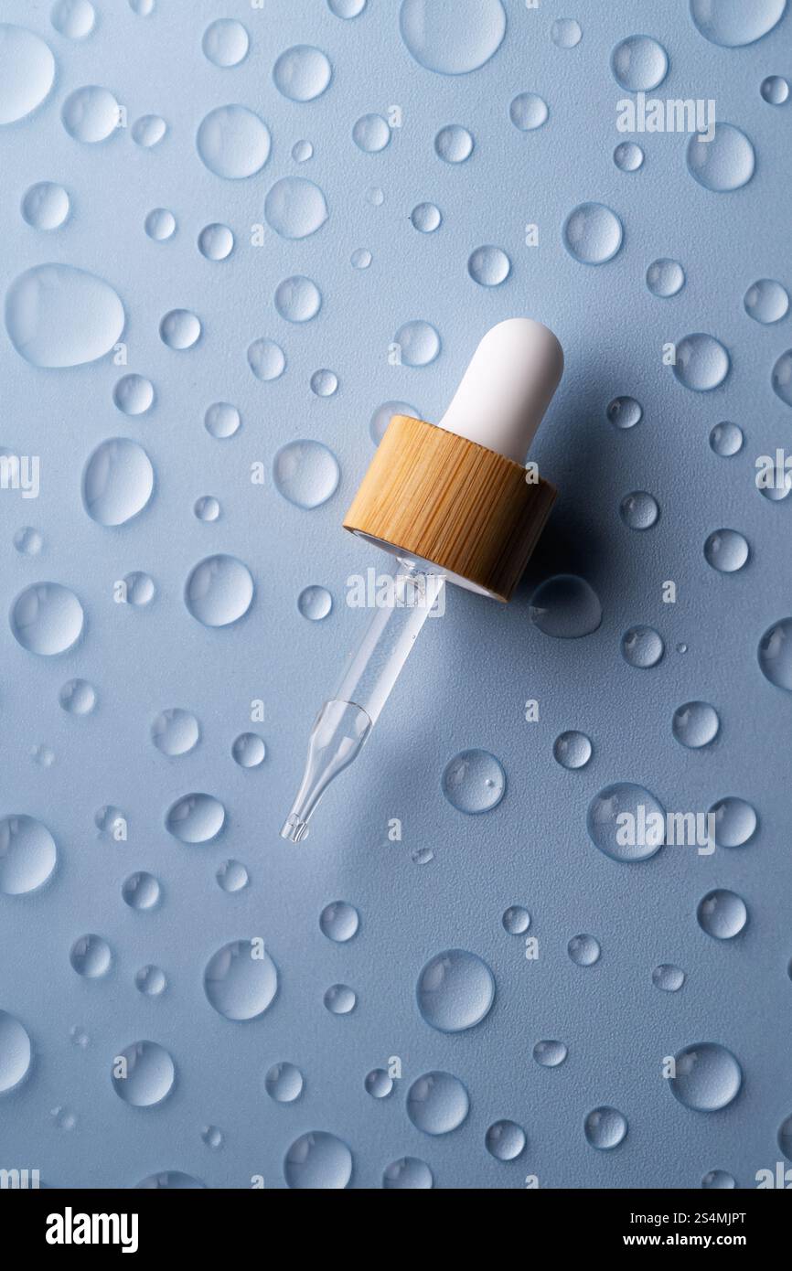 A glass pipette bottle lies on a textured blue surface Water droplets surround the bottle ...