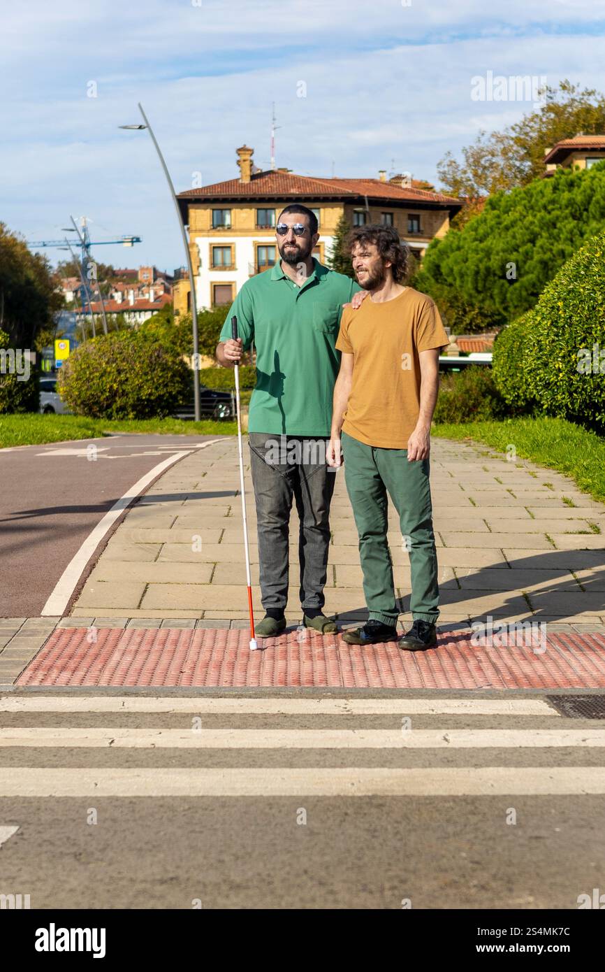 A blind man with a cane stands at a crosswalk, accompanied by his ...