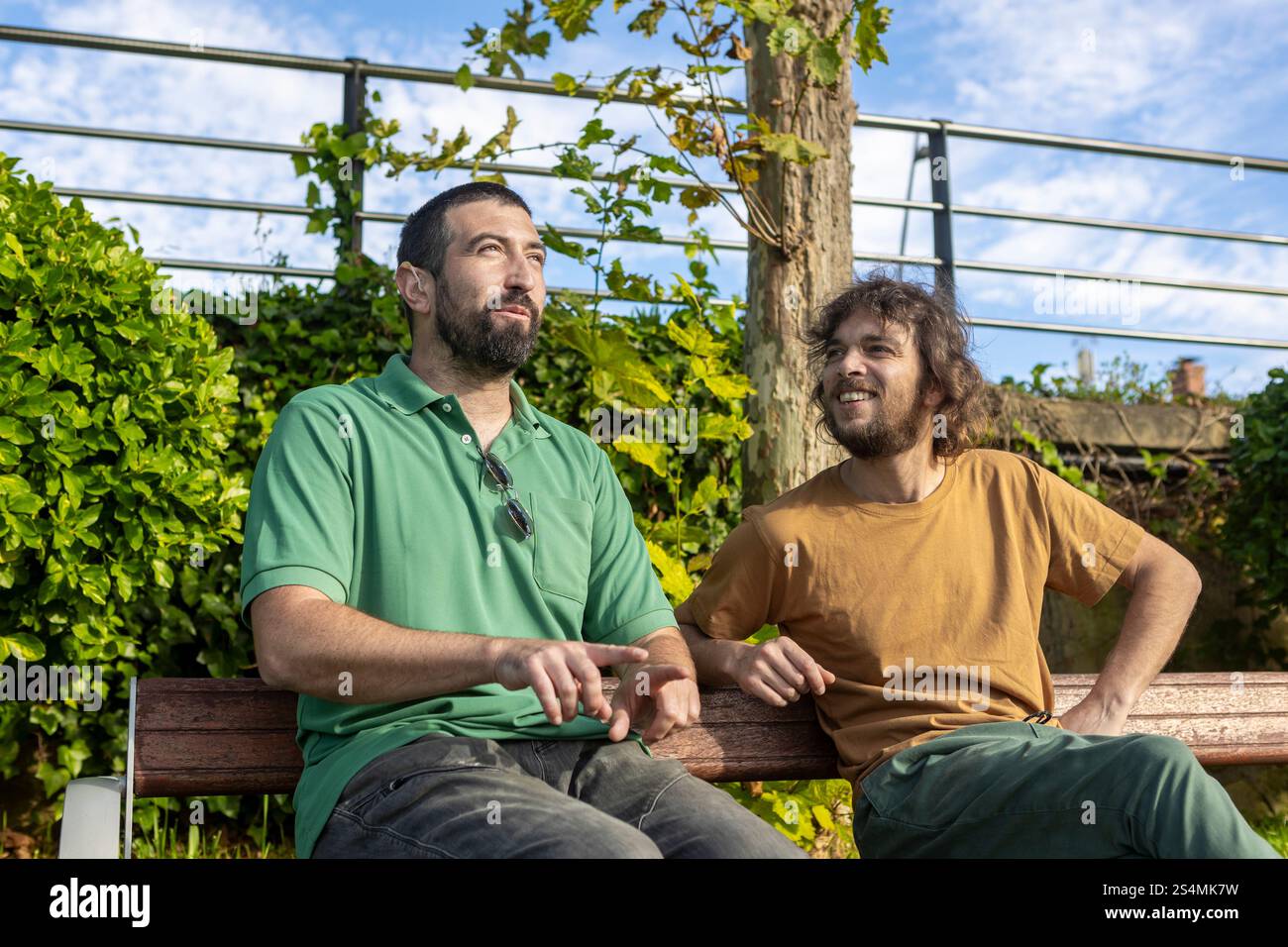 Two men sit on a bench surrounded by lush greenery One man, who is ...