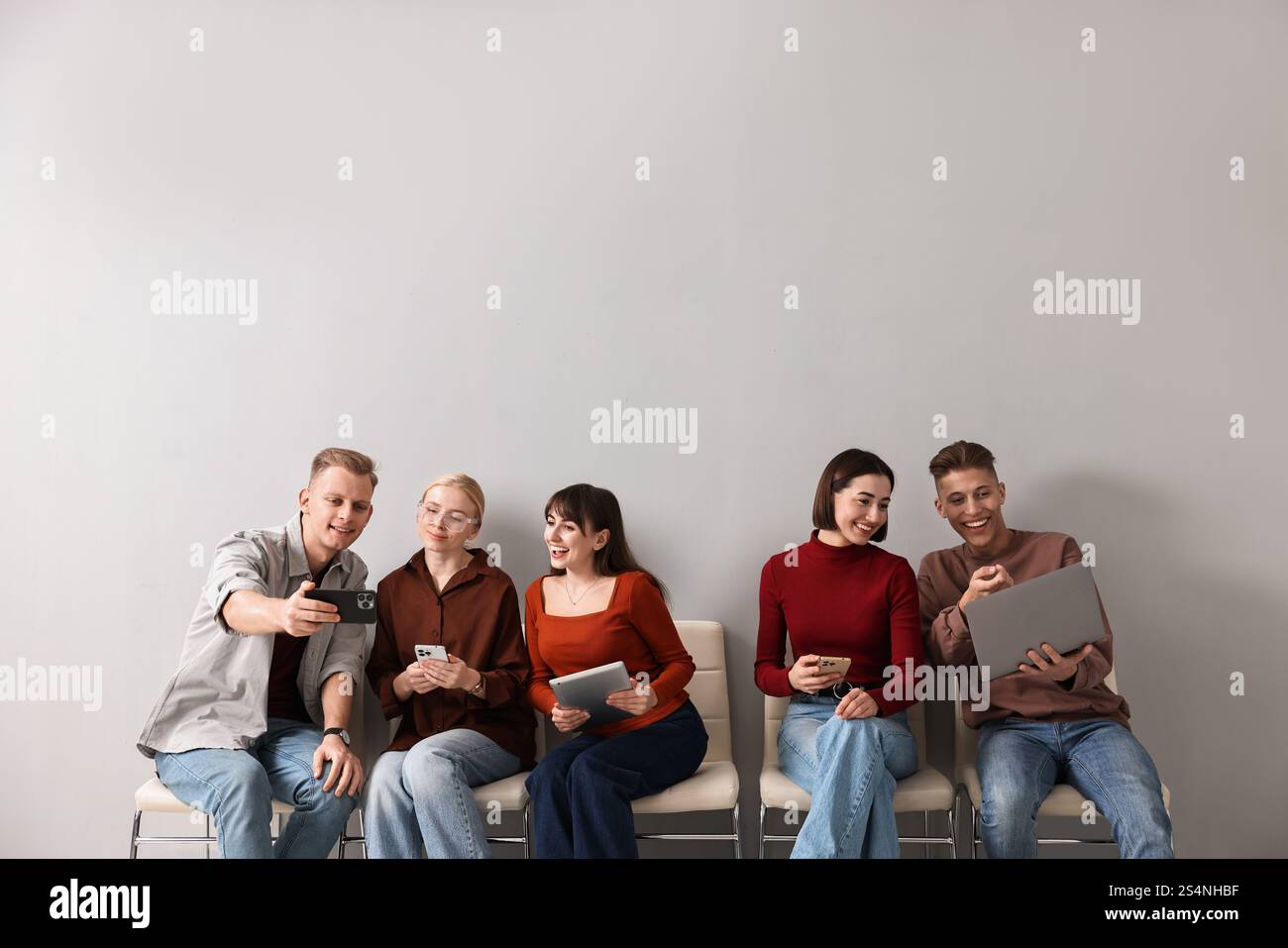 Group of people using different gadgets on chairs near light grey wall ...