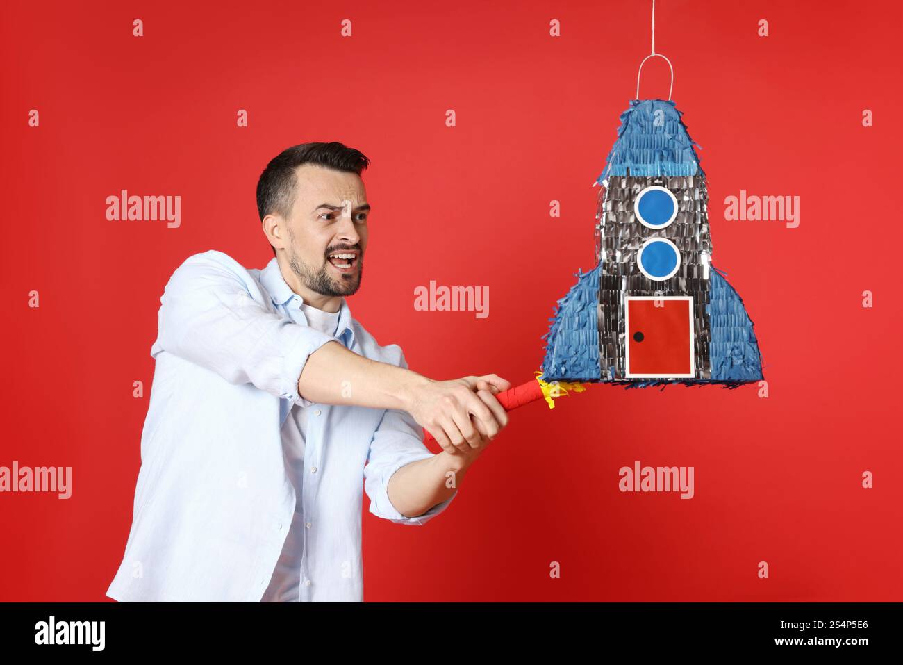 Emotional man hitting rocket shaped pinata with stick on red background ...