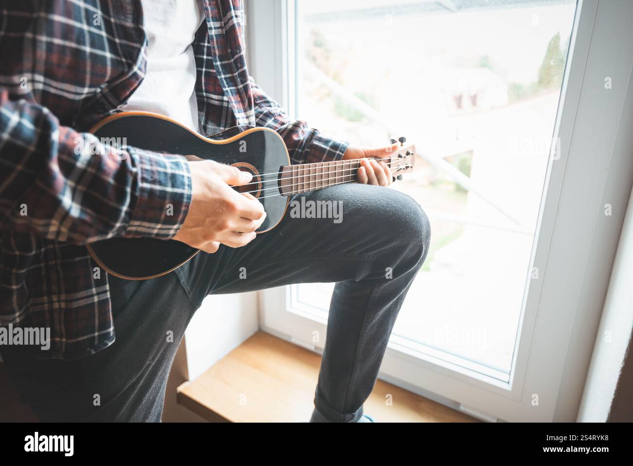 Man sits by a bright window, holding a glossy black ukulele. With ...