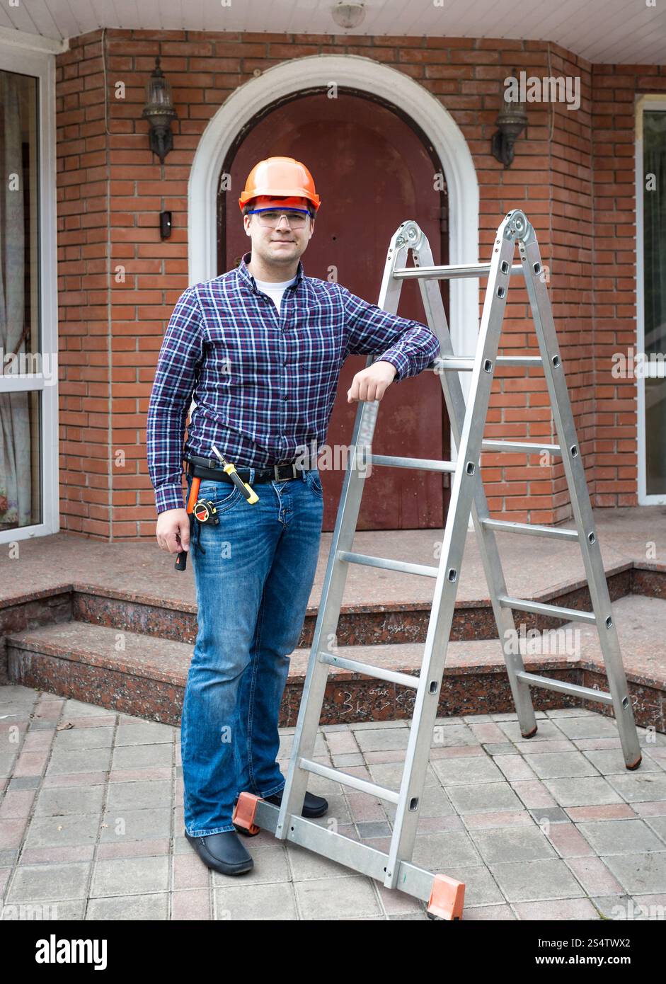 Male builder in helmet posing with metal ladder against house entrance ...