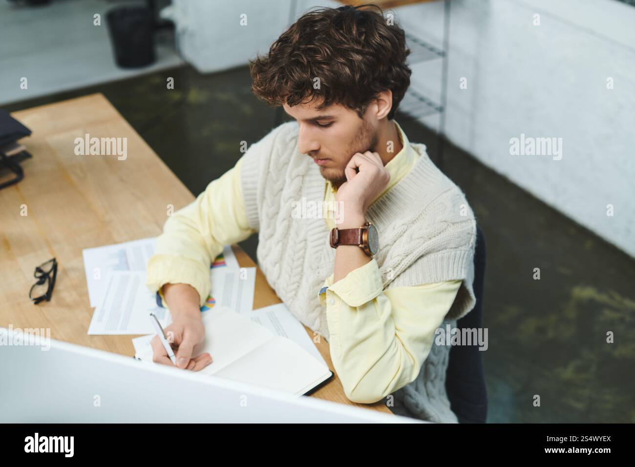 A young man in a sweater vest studies his notes at a modern office desk ...