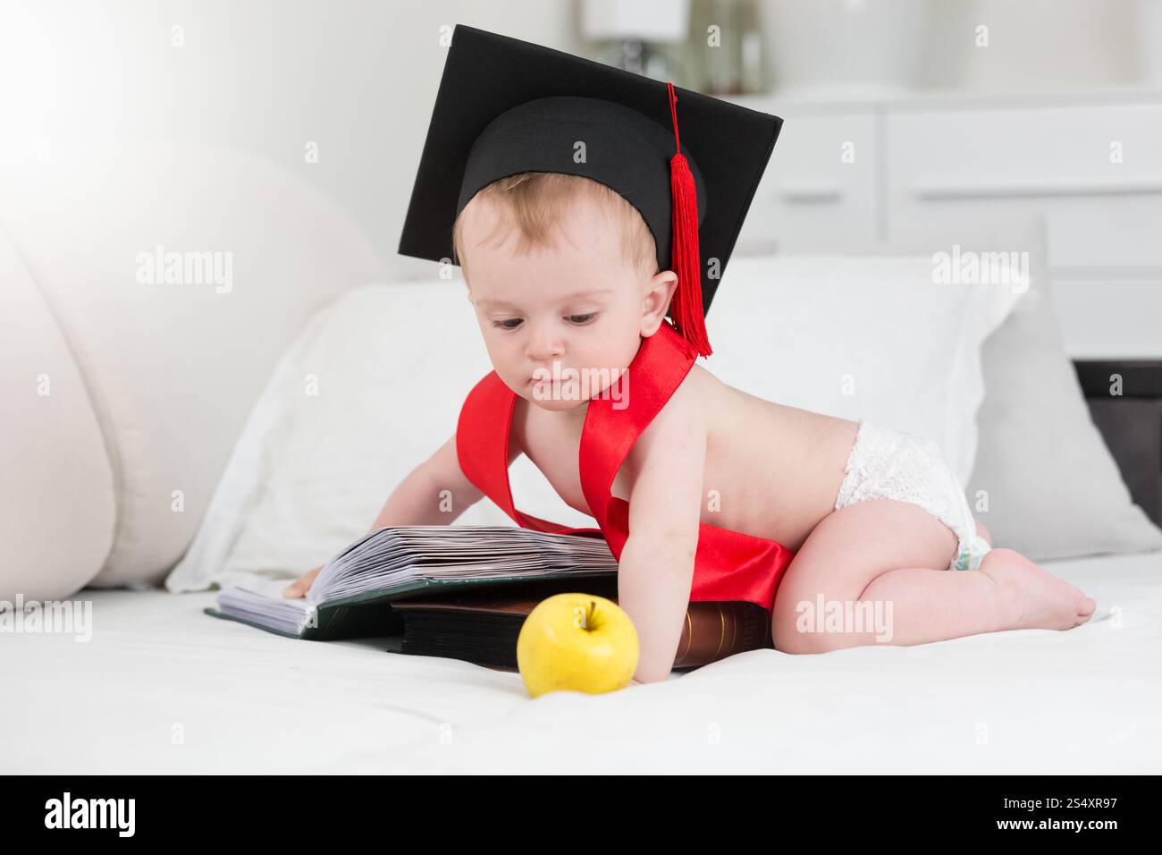 Adorable baby boy in graduation cap posing with apple and big book ...