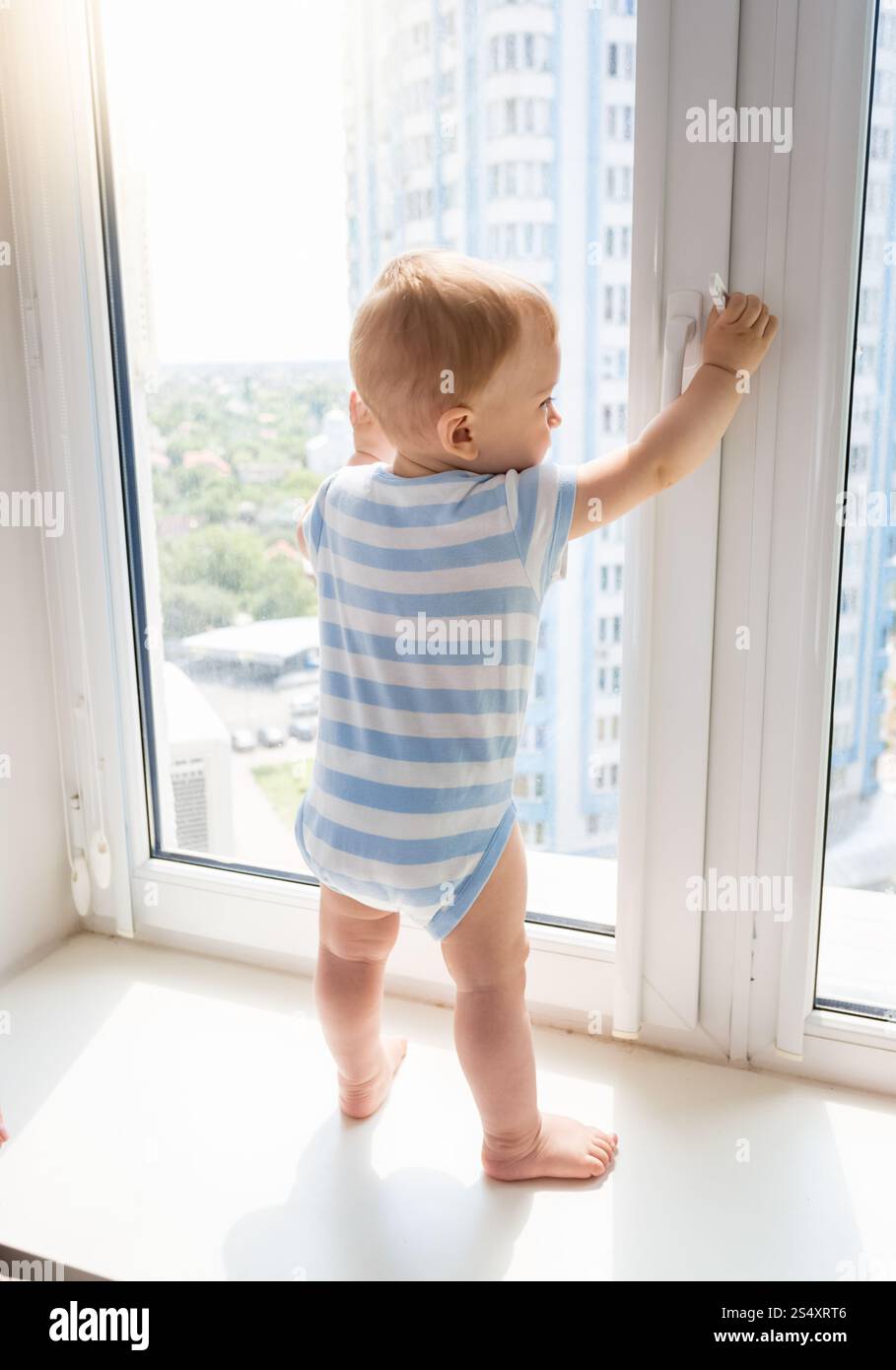 Little baby boy standing on window sill and pulling window handle ...