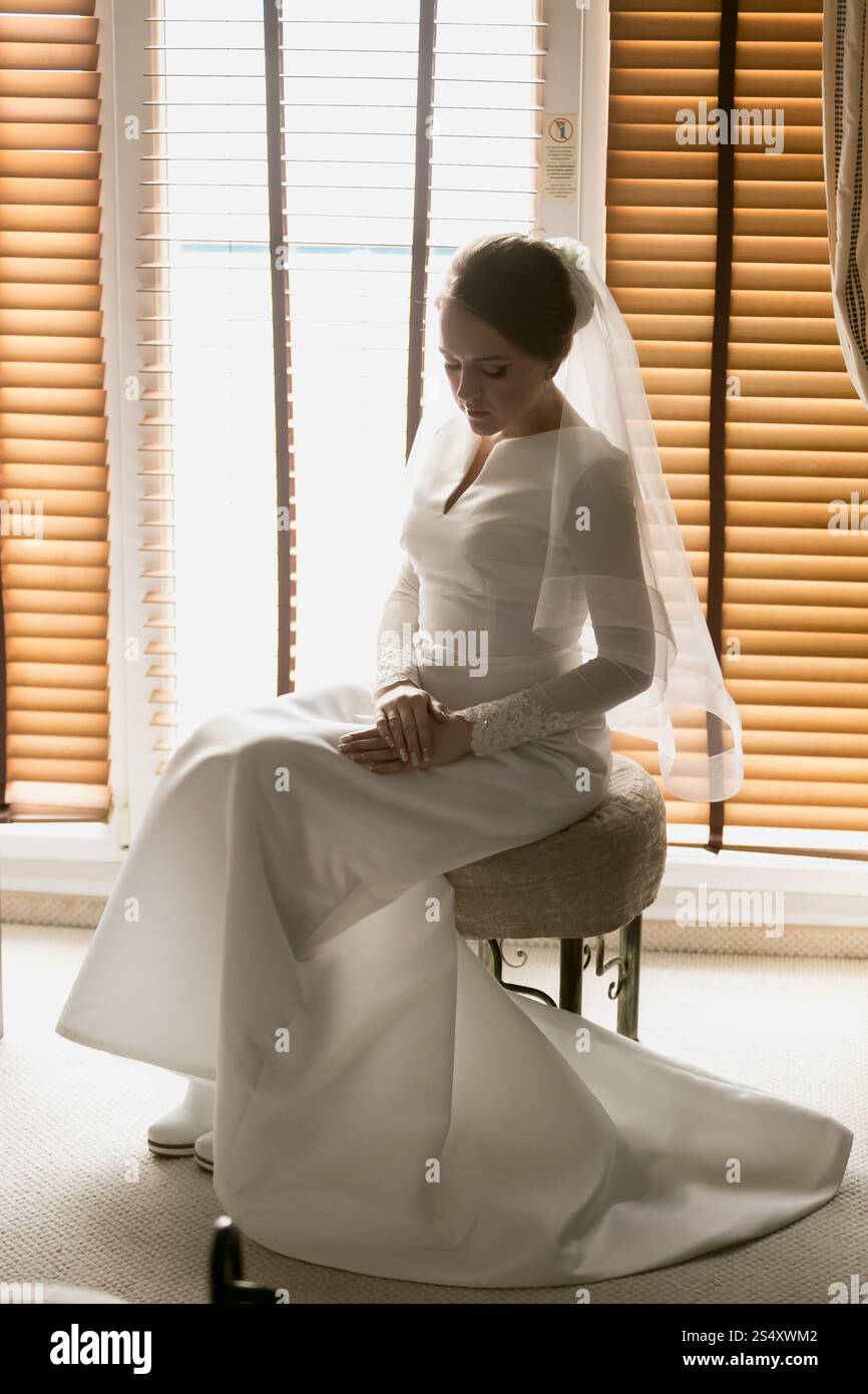 Beautiful young bride posing on chair against big window at hotel room ...