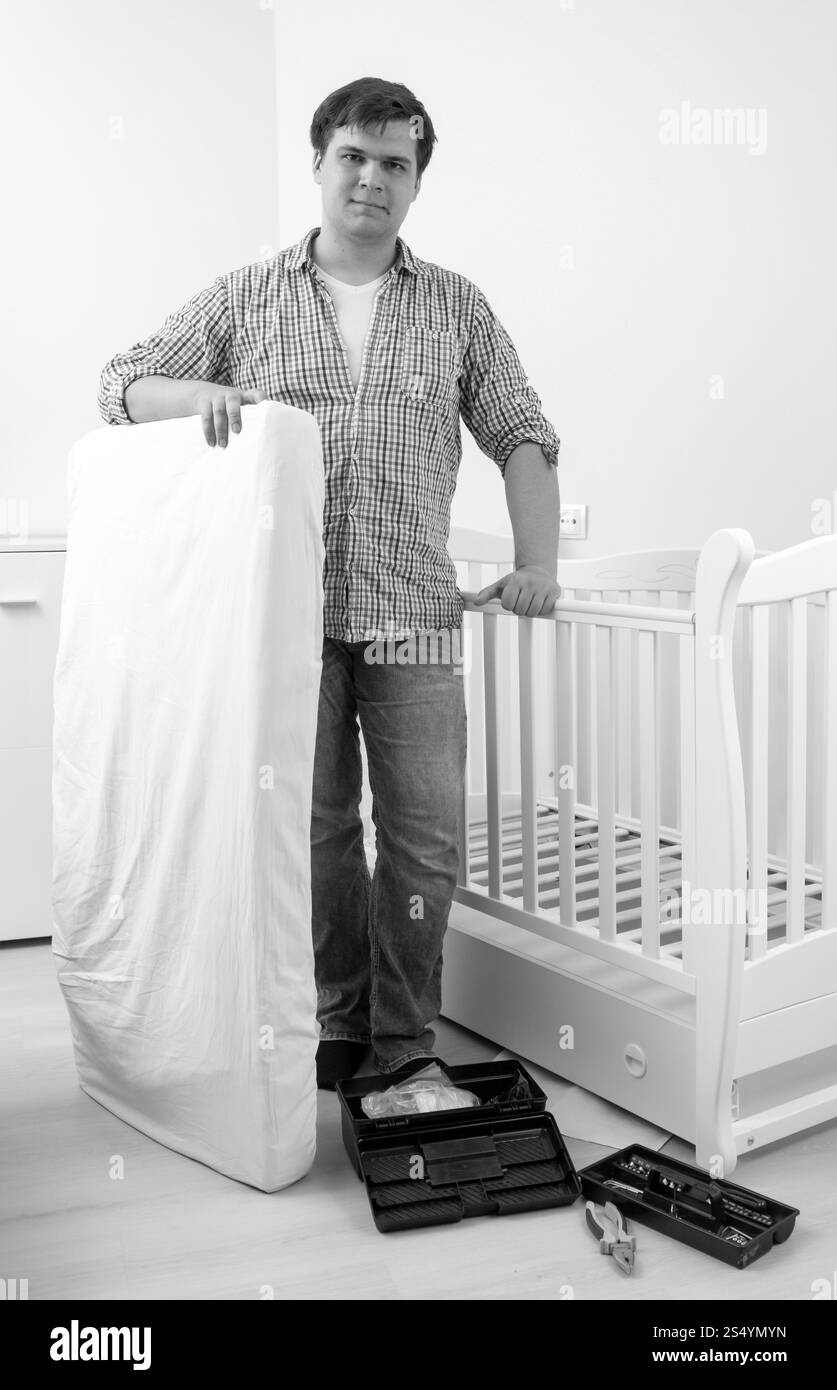 Black and white portrait of young handsome man posing at babys cot with ...