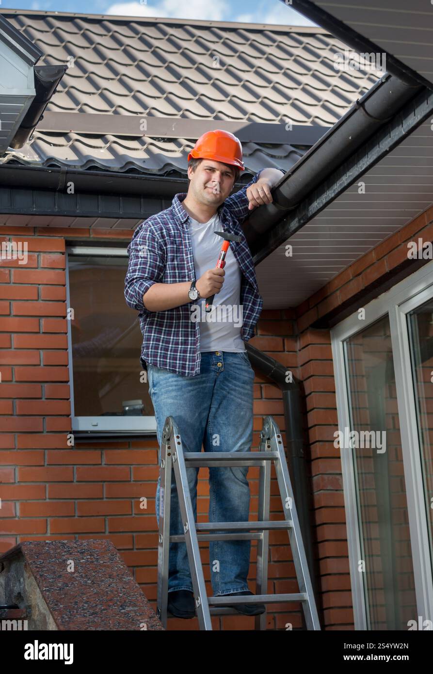 Smiling carpenter posing with hammer on step ladder under the roof ...