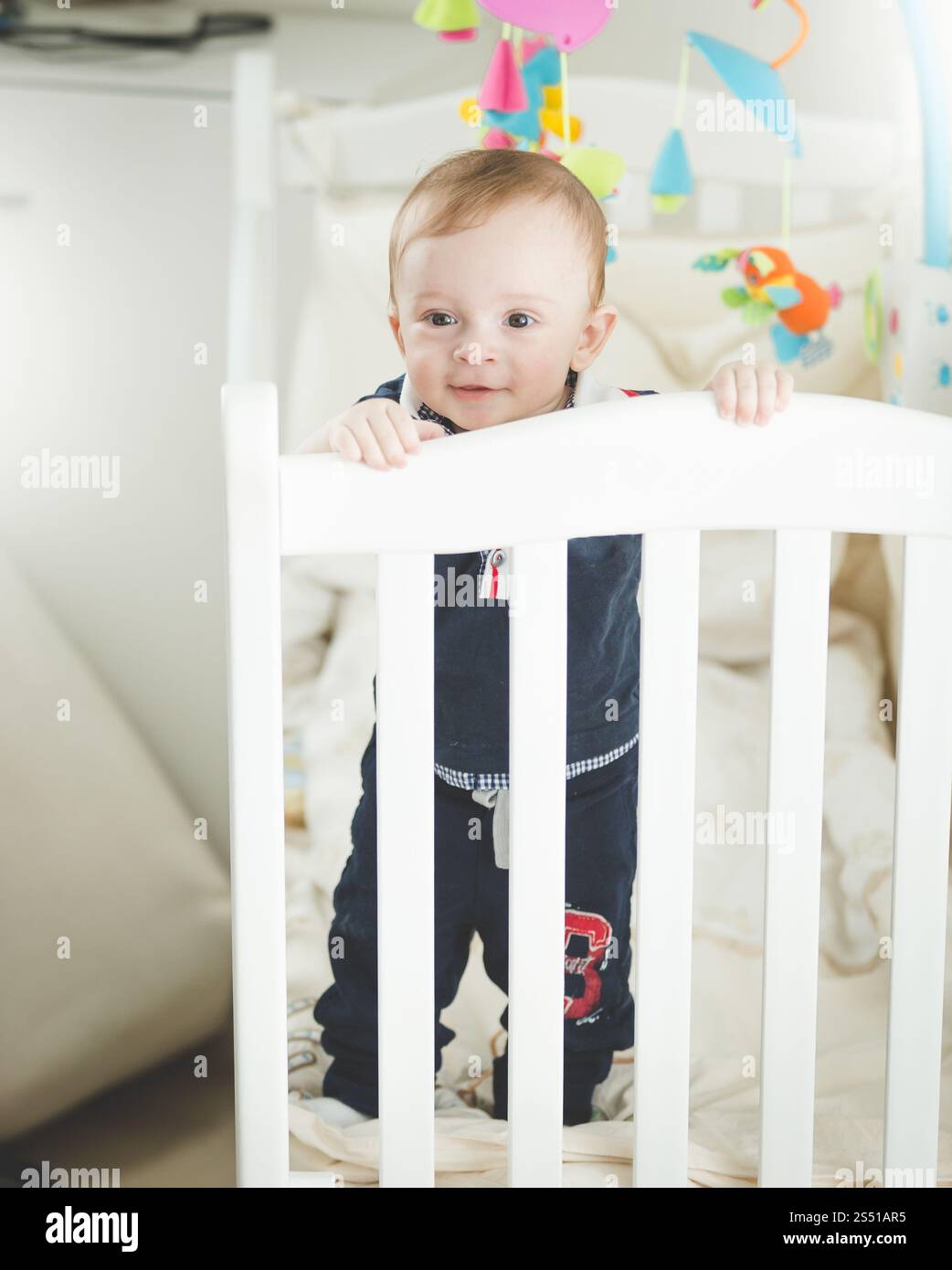 Portrait of adroable 1 year old baby boy standing in white wooden crib ...