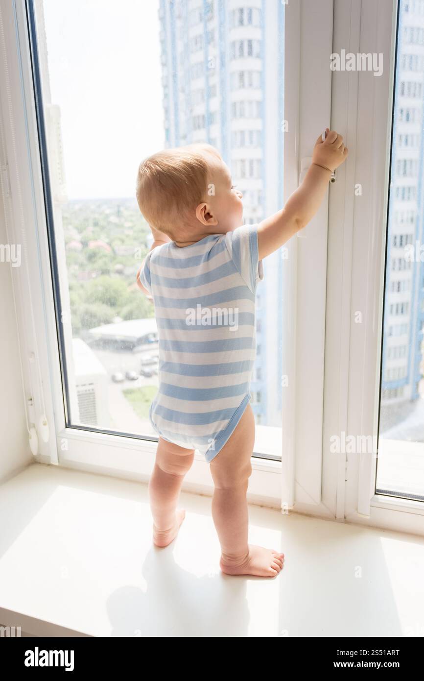 9 months old baby standing on windowsil and trying to open the lock on ...