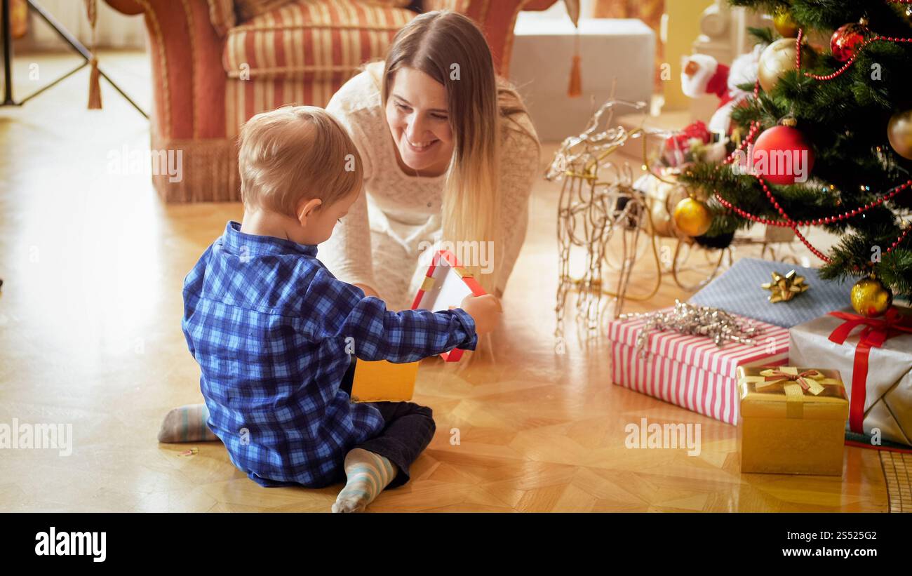 Boy sitting on floor with mother ond opening box with Christmas gift ...