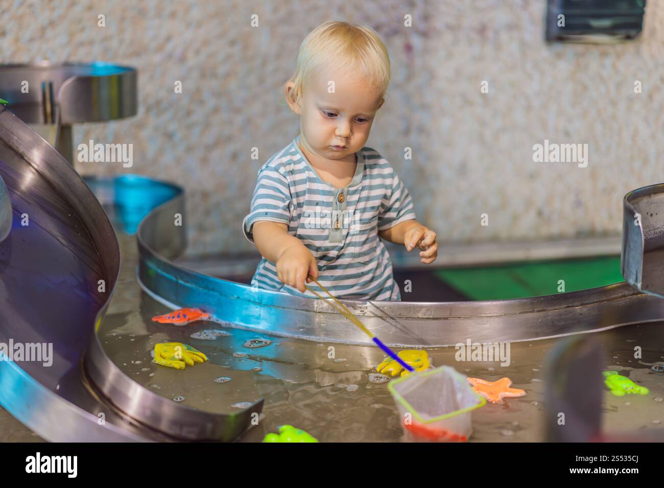 Toddler boy catching toy fish with a net in a vibrant water play area ...
