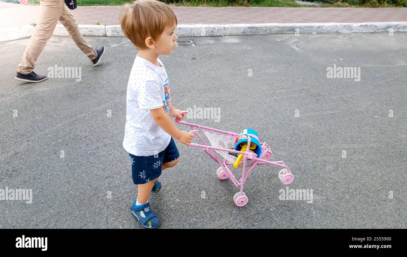 Little boy walking on road and pushing toy stroller for dolls. Little ...
