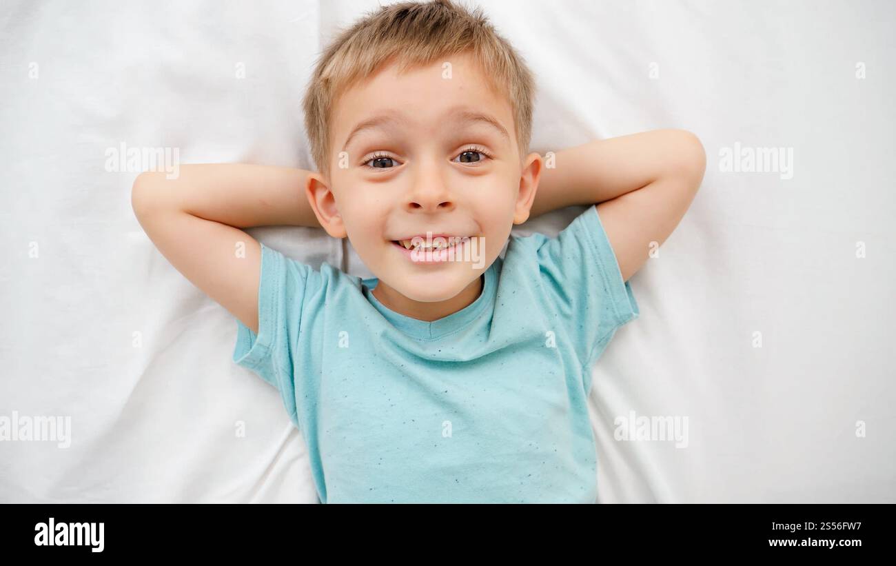 Portrait of smiling little boy in pajamas lying on big bed with white ...