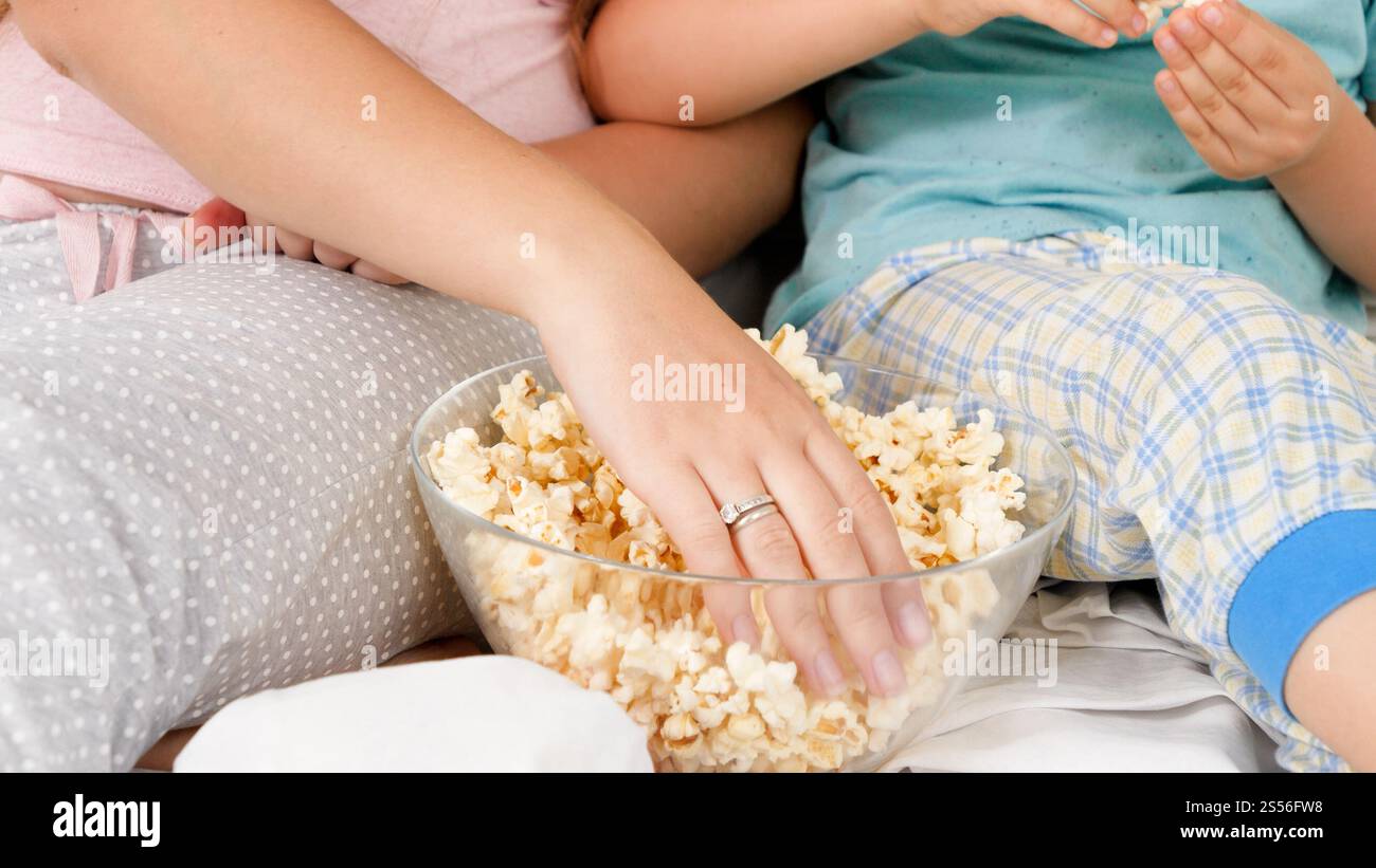 Closeup of mother and son in pajamas taking popcorn from big glass bowl ...