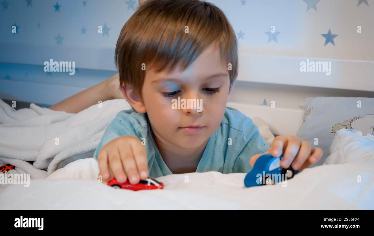 Smiling little boy playing with two toy cars while lying in bed at ...