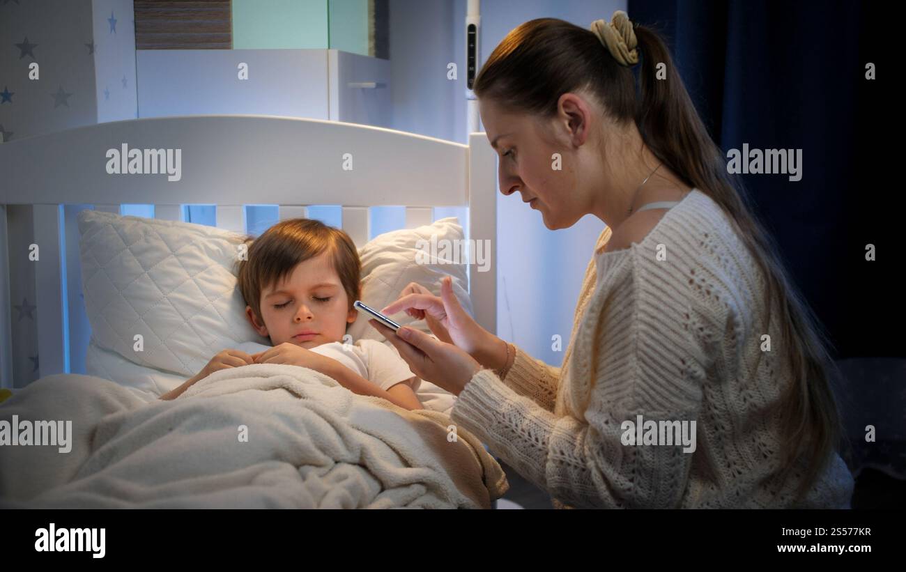 Young mother sitting next to her sick child lying in bed and browsing for medicines on ...
