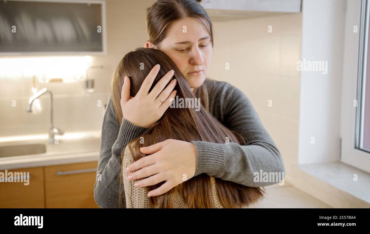 Young mother hugging and consoling upset crying daughter on kitchen. Parent supporting and ...