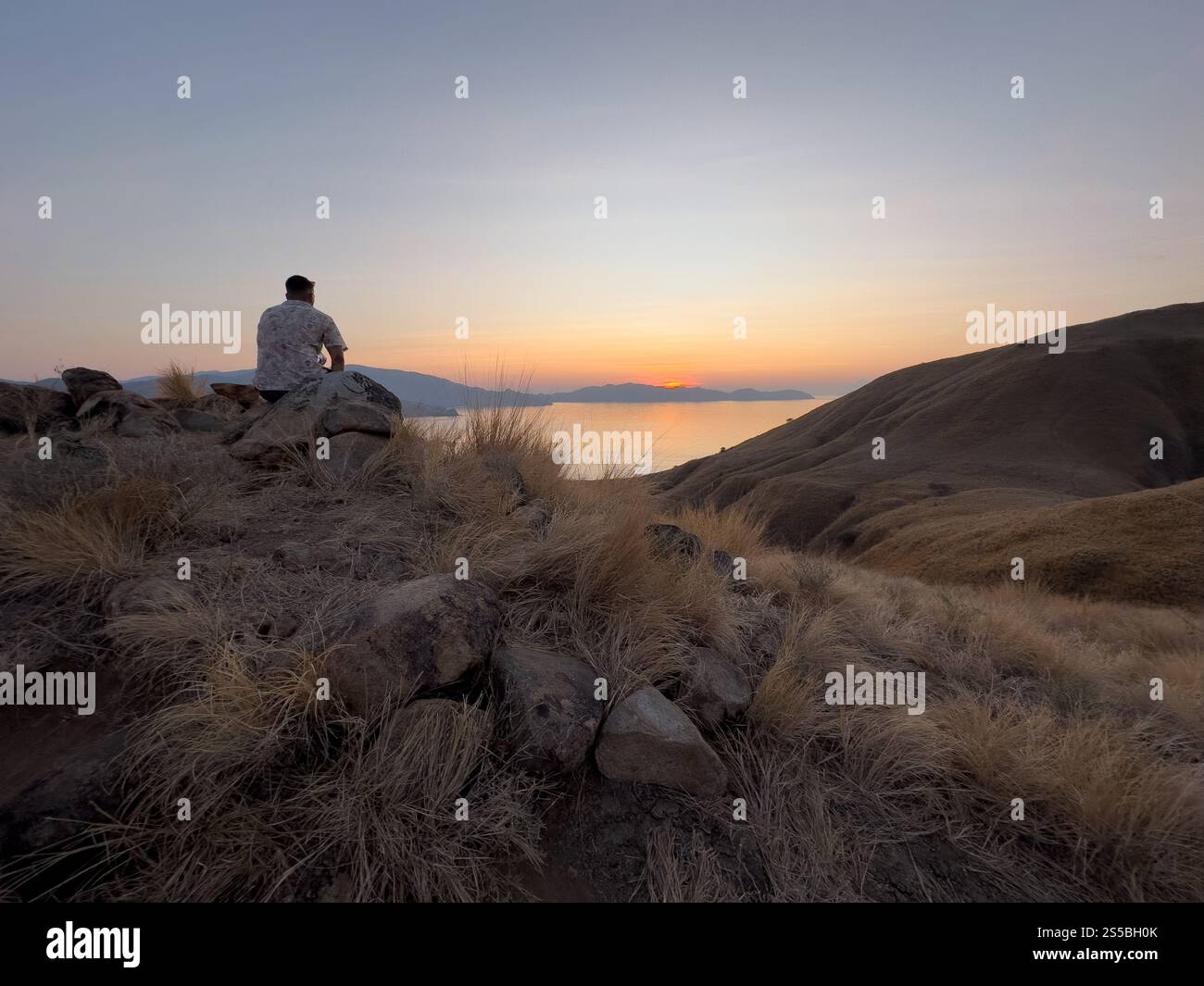 Rear view of a man sitting by the ocean watching the sunset over Lawa ...