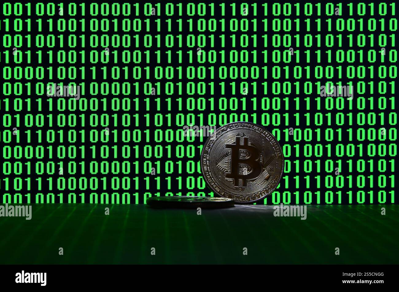 A pair of bitcoins lies on a cardboard surface on the background of a monitor depicting a binary ...