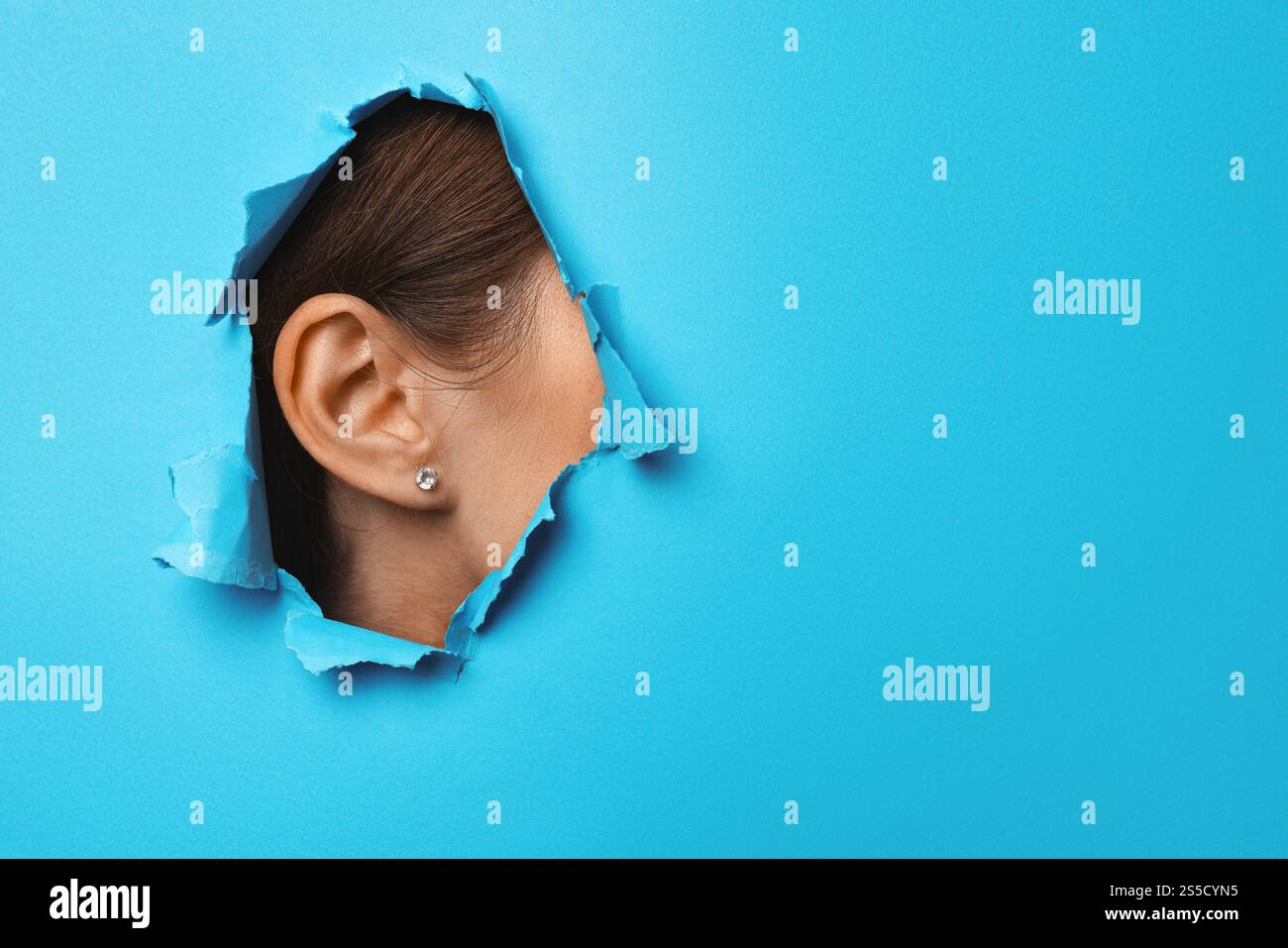 Woman showing her ear through hole in light blue paper, closeup. Space ...