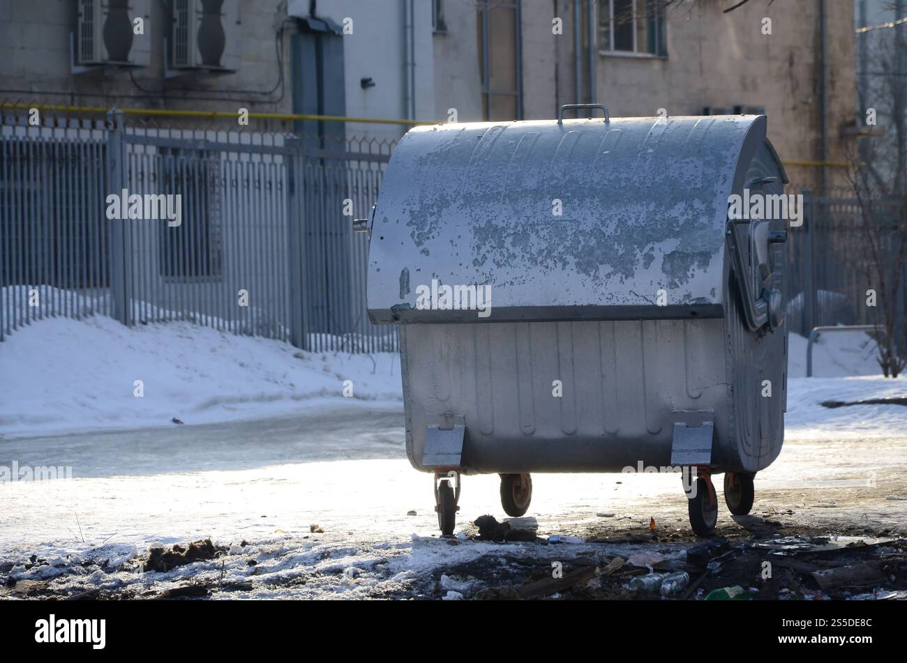 A silver garbage container stands near residential buildings in winter ...