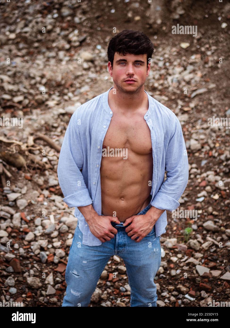 A muscular man stands amidst rocks, wearing a light shirt and jeans. He ...