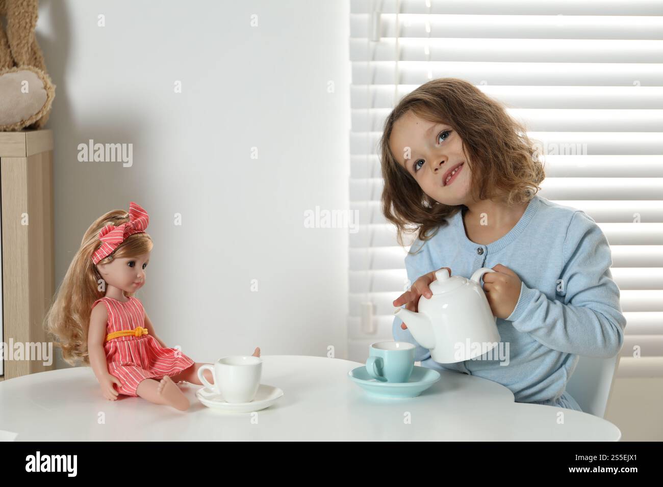 Cute little girl playing tea party with doll at white table in room ...
