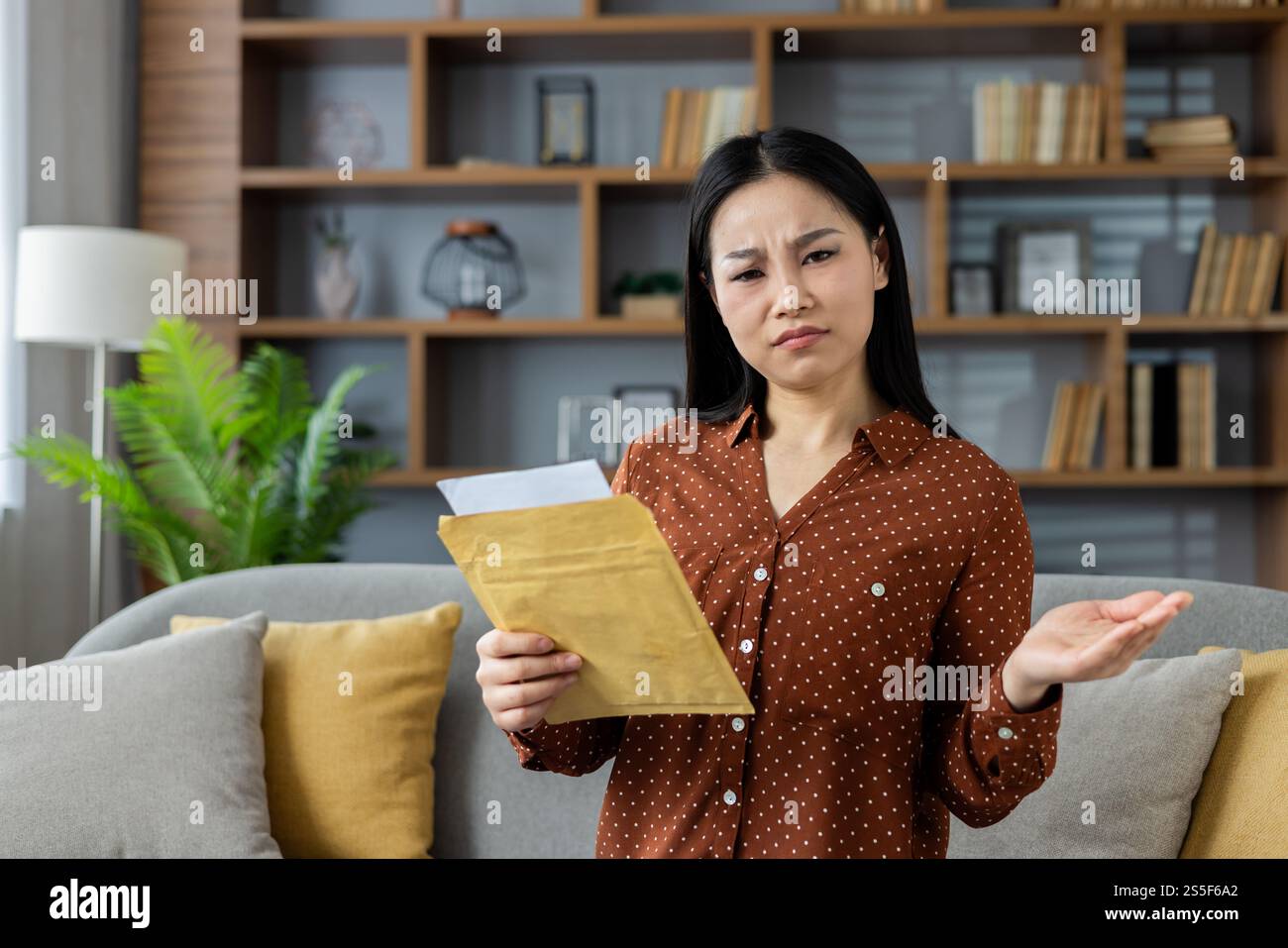 Asian woman with perplexed look holds envelope, expressing confusion ...