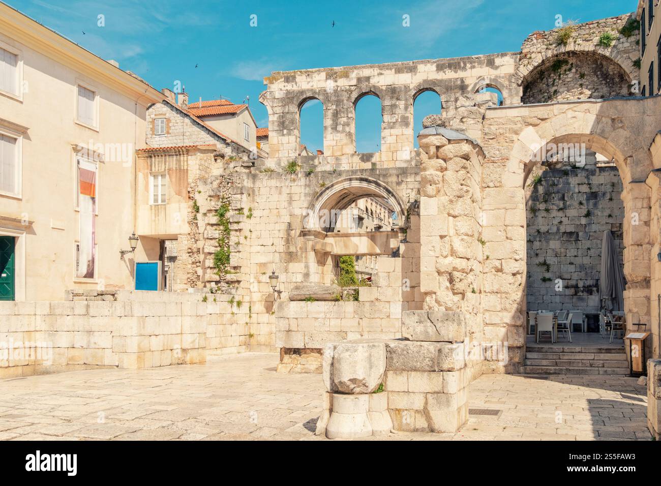 Ancient stone ruins with arches and columns in a European city square under a clear blue sky ...
