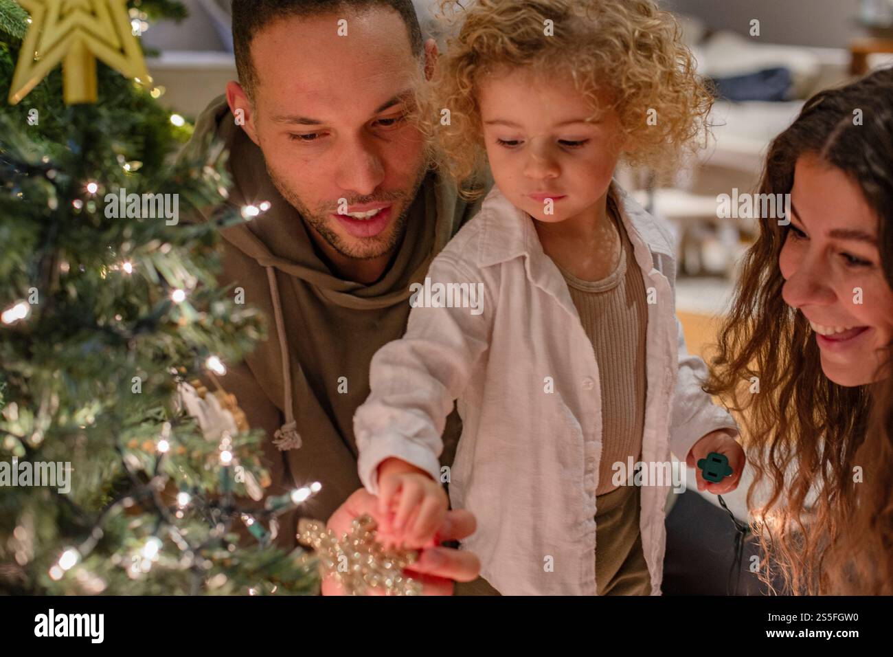 A family of three is decorating a Christmas tree, with a toddler ...
