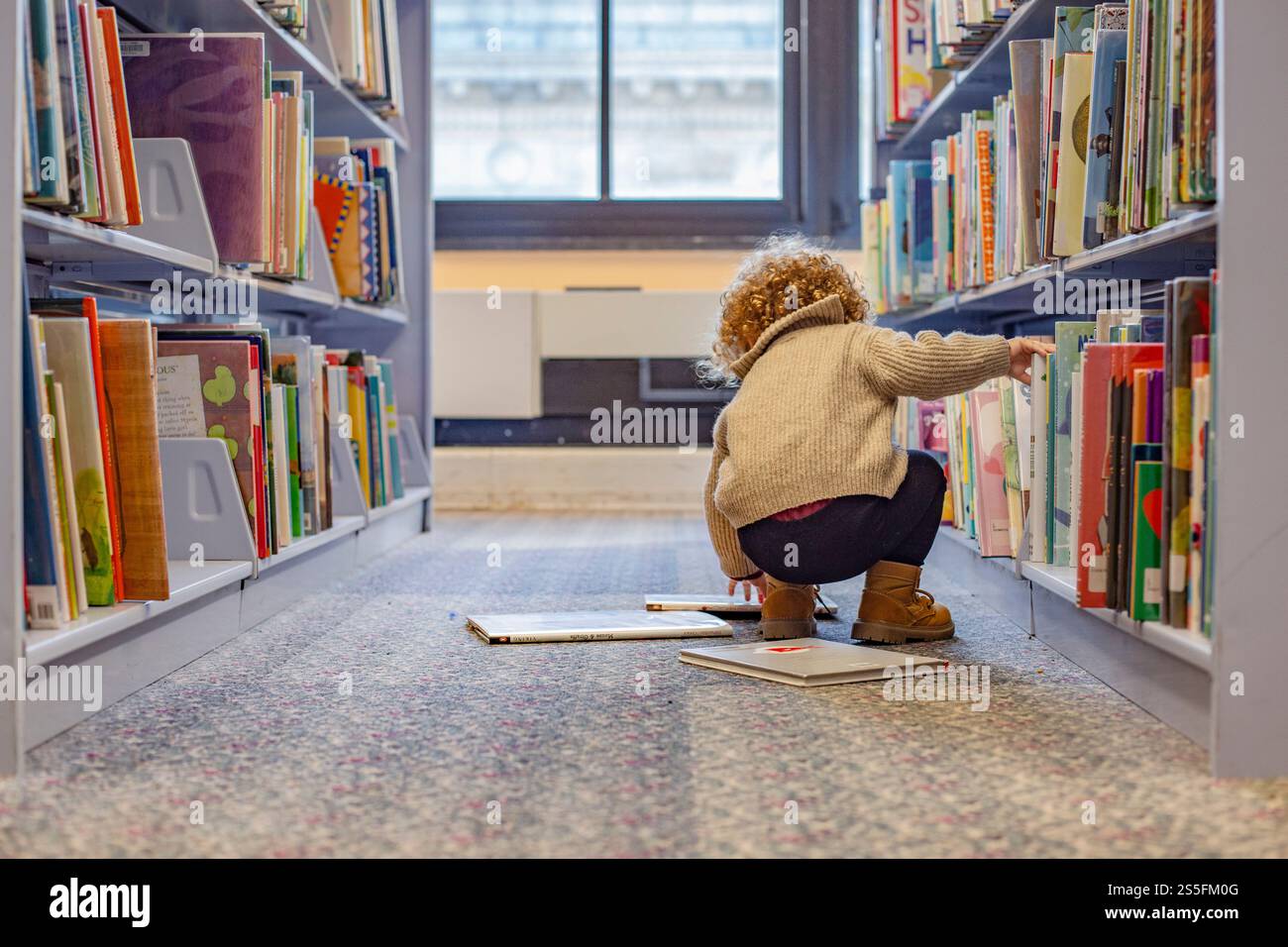 Toddler selecting books from a library shelf with books on the floor ...
